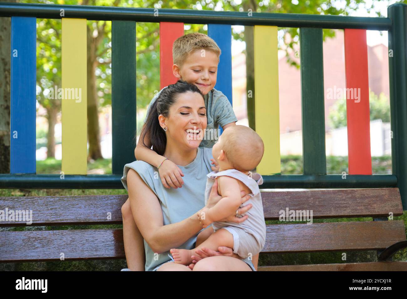 A joyful mother sits on a park bench, cuddling her infant and smiling ...