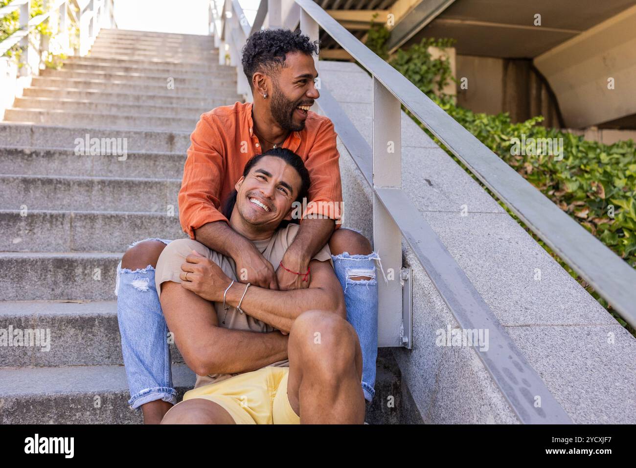 An affectionate and joyful gay couple share a laugh while embracing on outdoor city steps ...