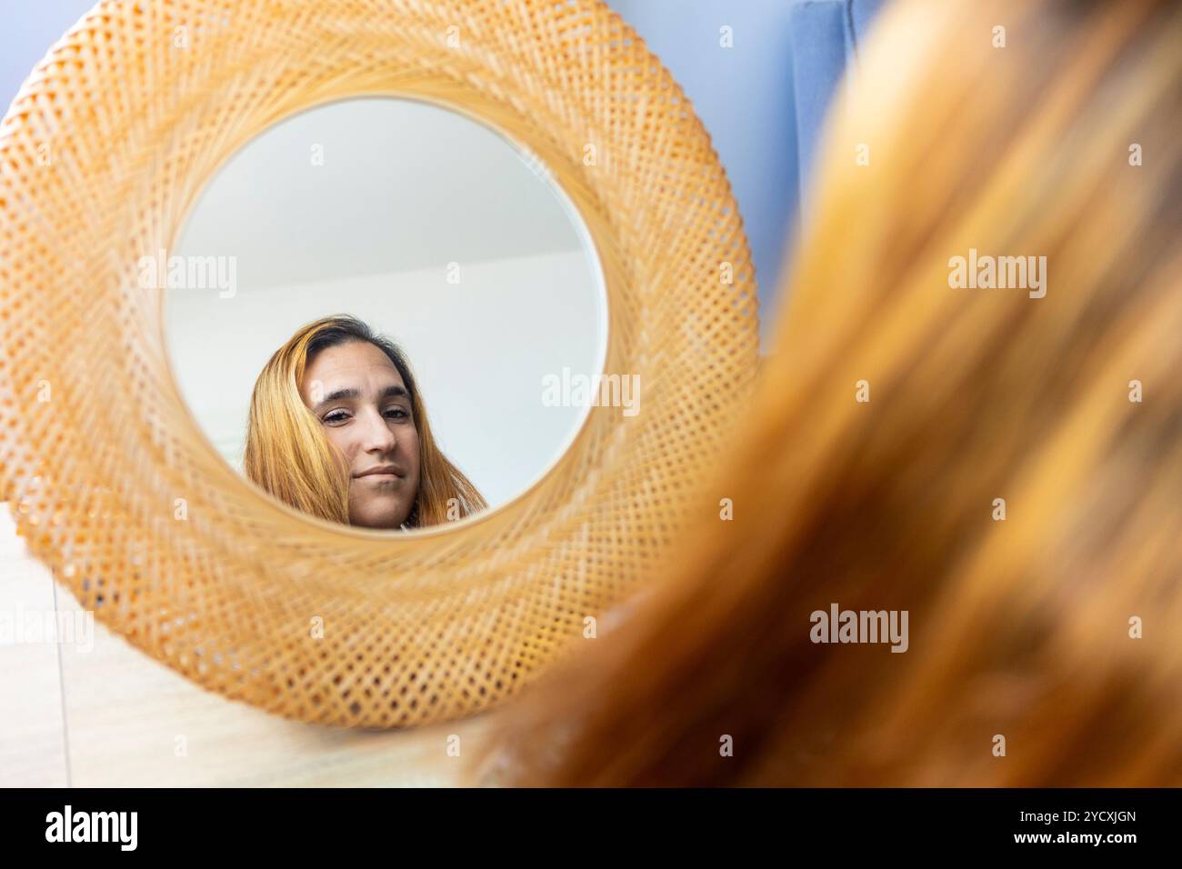 A trans woman looks in a round mirror, her expression soft and ...