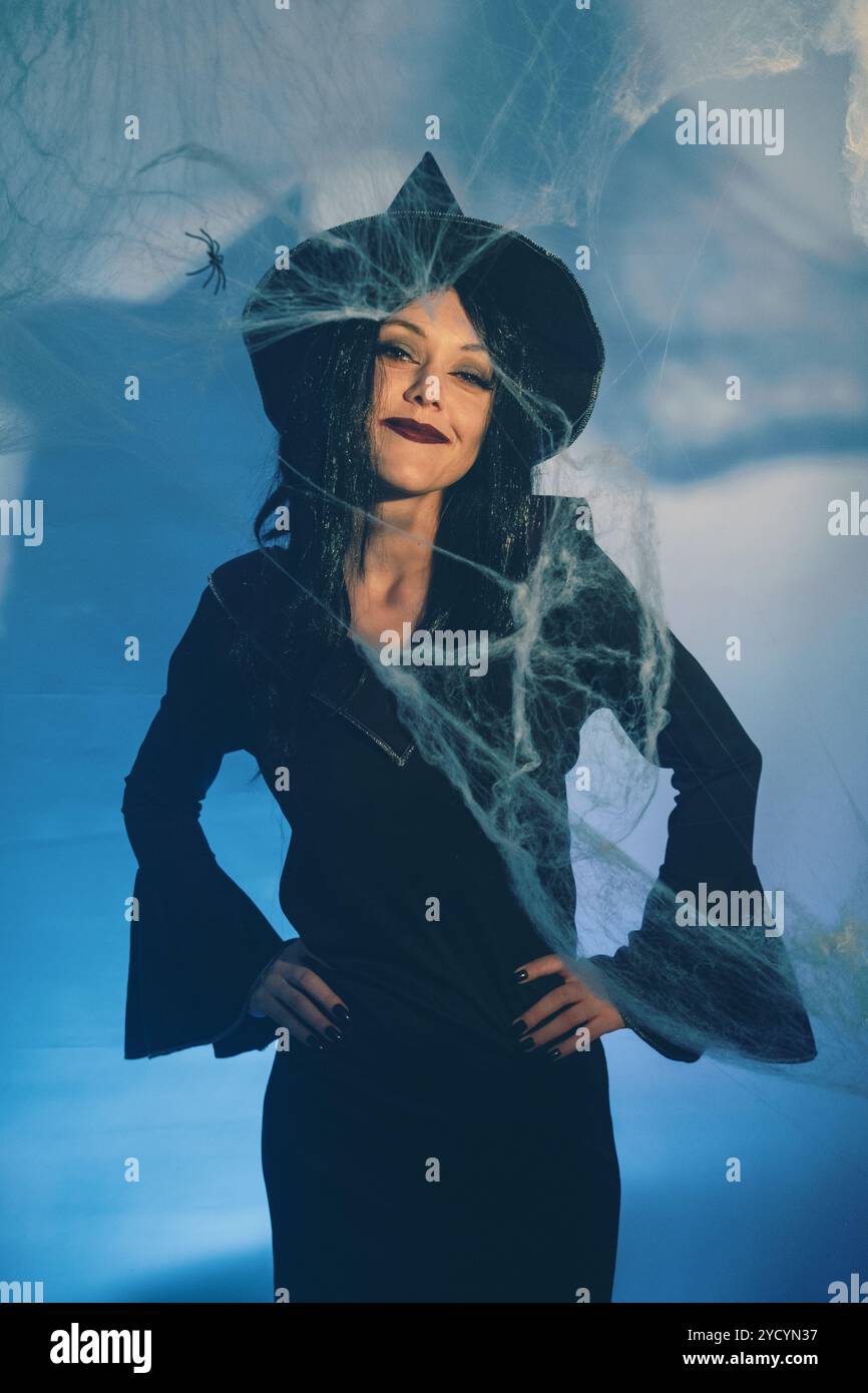 A charming witch poses in front of spider webs, her hands raised in a ...