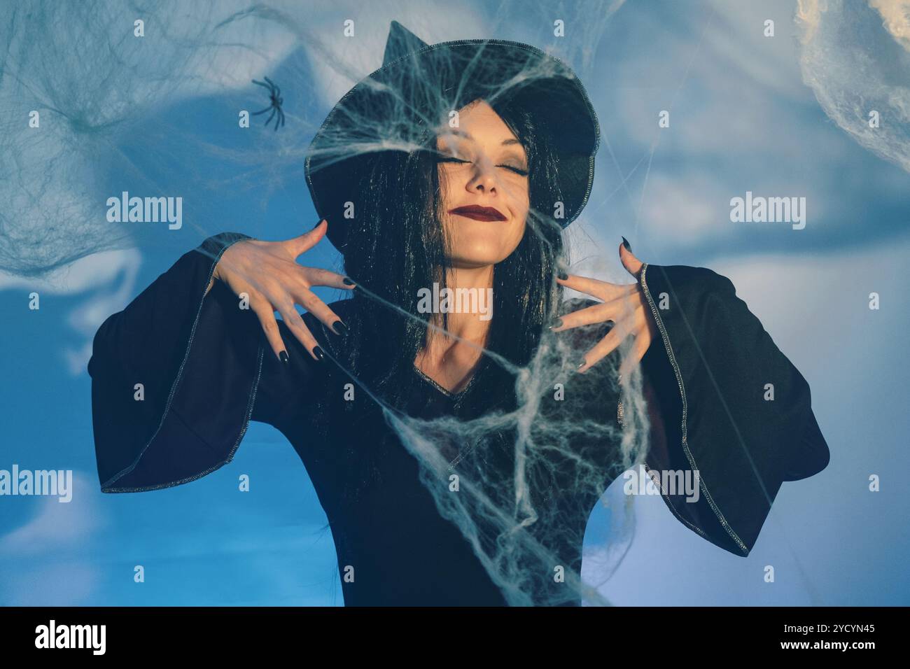 A charming witch poses in front of spider webs, her hands raised in a ...