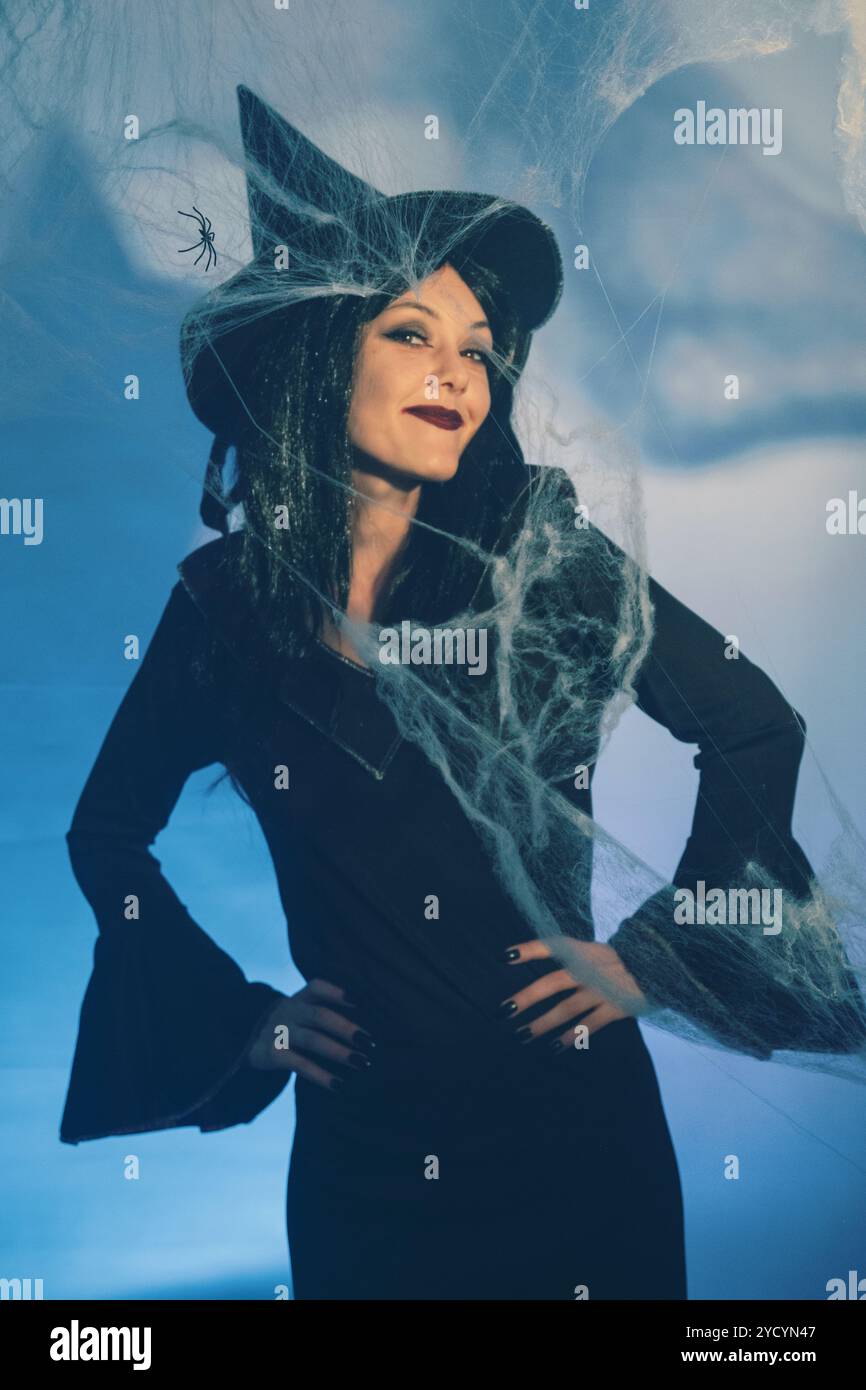 A charming witch poses in front of spider webs, her hands raised in a ...