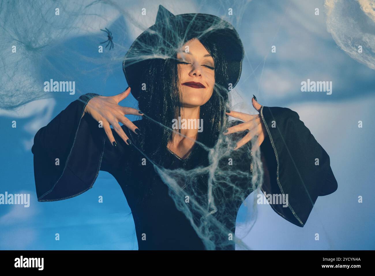 A charming witch poses in front of spider webs, her hands raised in a ...