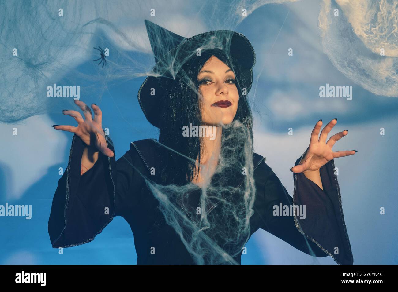 A charming witch poses in front of spider webs, her hands raised in a ...