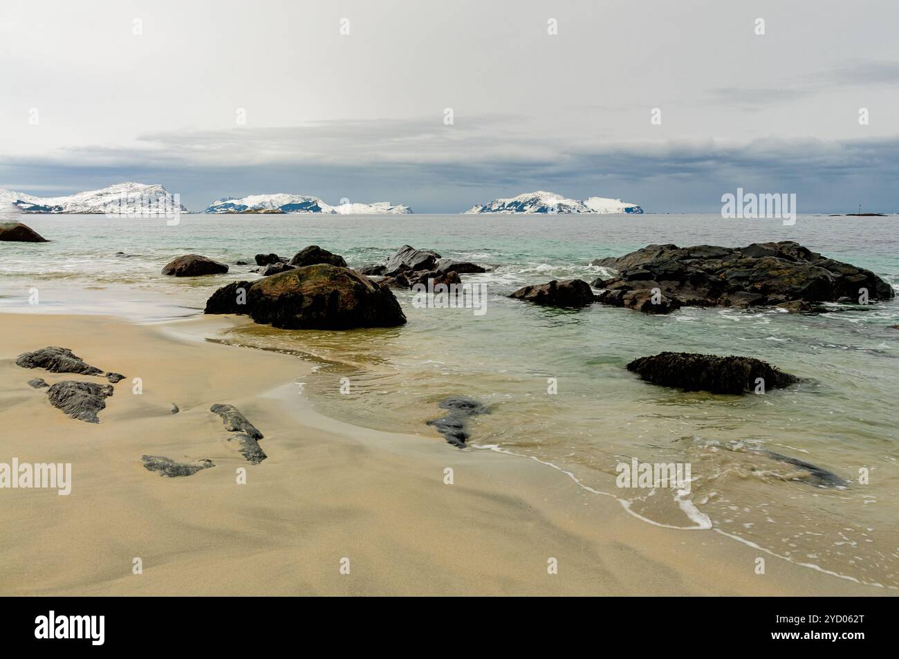 The image shows a sandy beach with large rocks and small waves. The ...