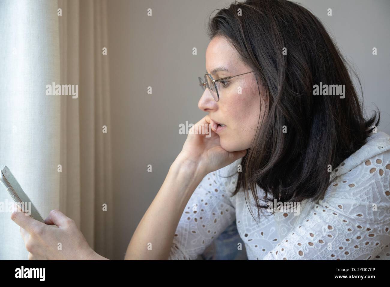 A woman in glasses looks intently at her smartphone with a thoughtful ...