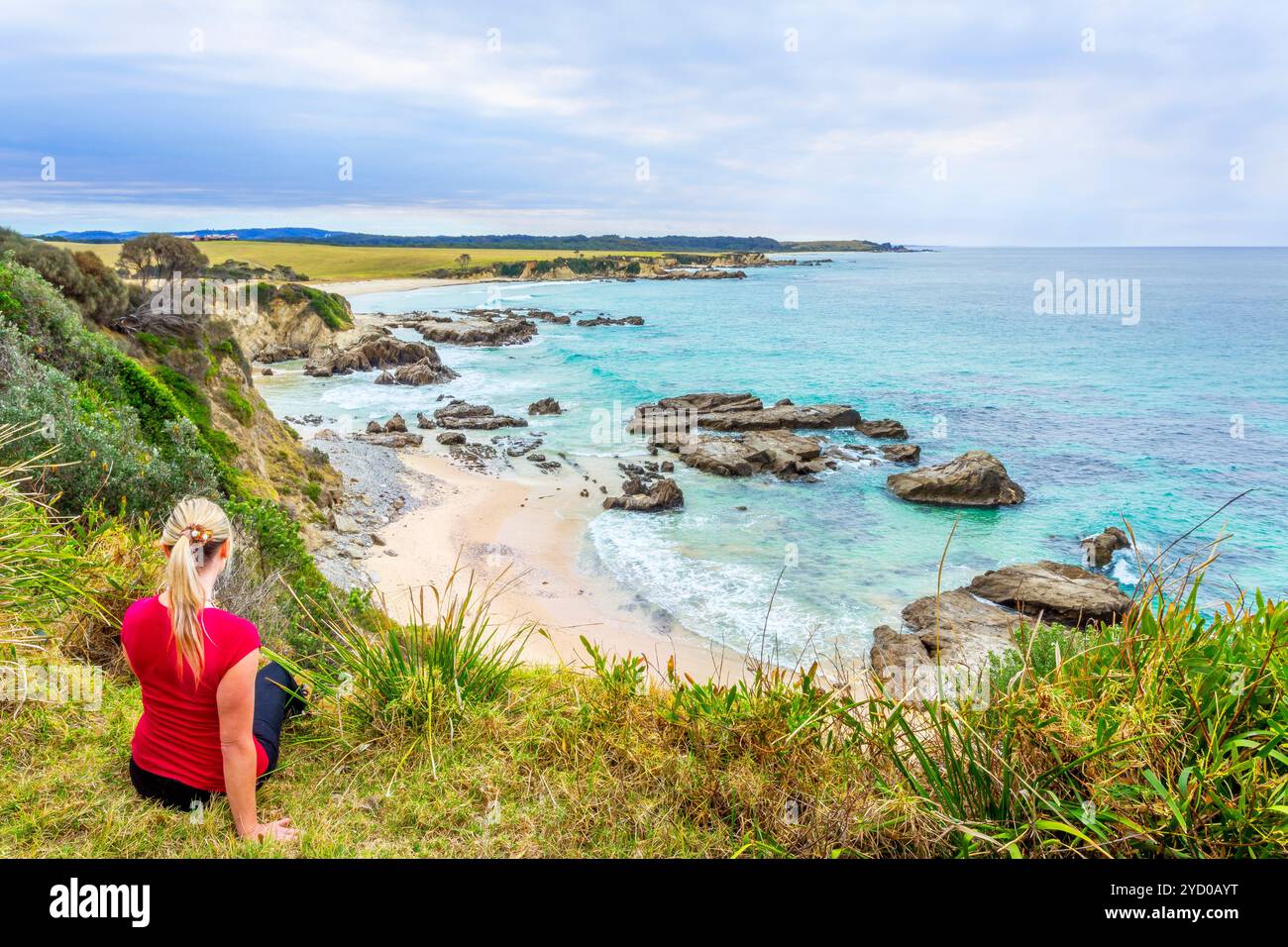 Female sitting on the coastal cliffs looking down onto the rocky beach ...