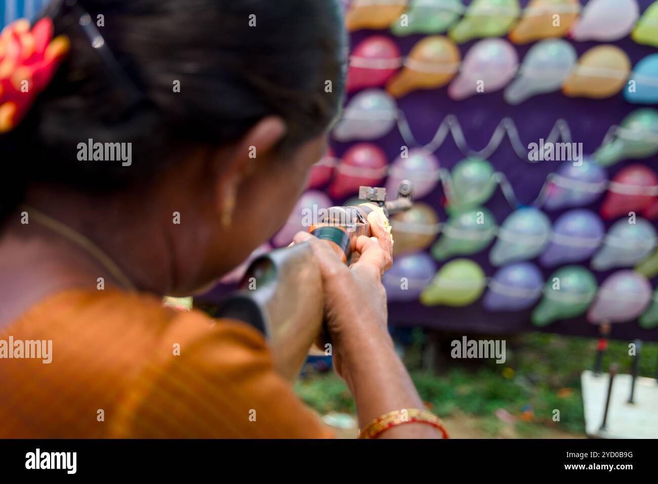 A Indian woman focuses intently, aiming an air rifle at a colorful ...