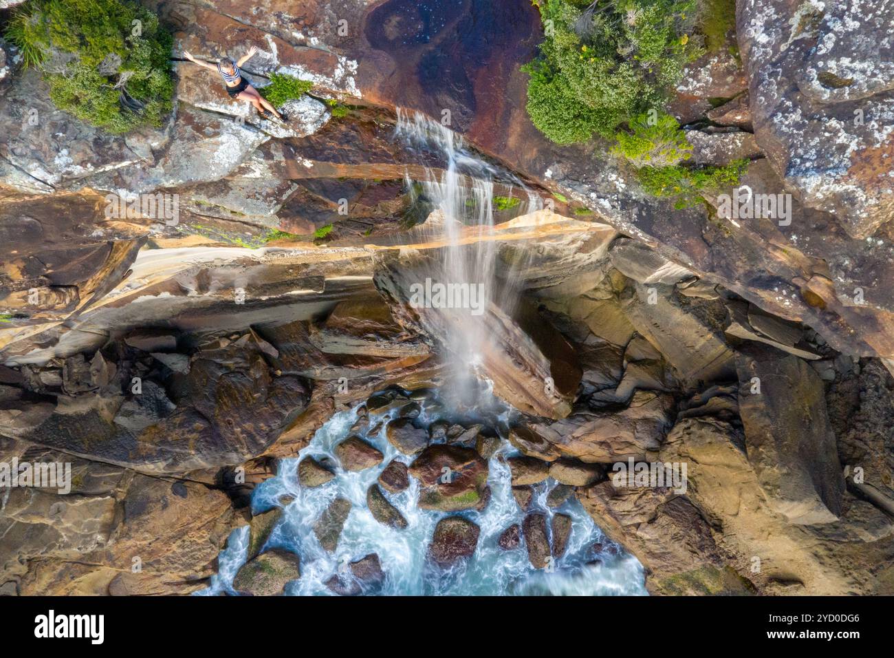 Clifftop woman sitting by the edge of a waterfall tumbling into ocean ...