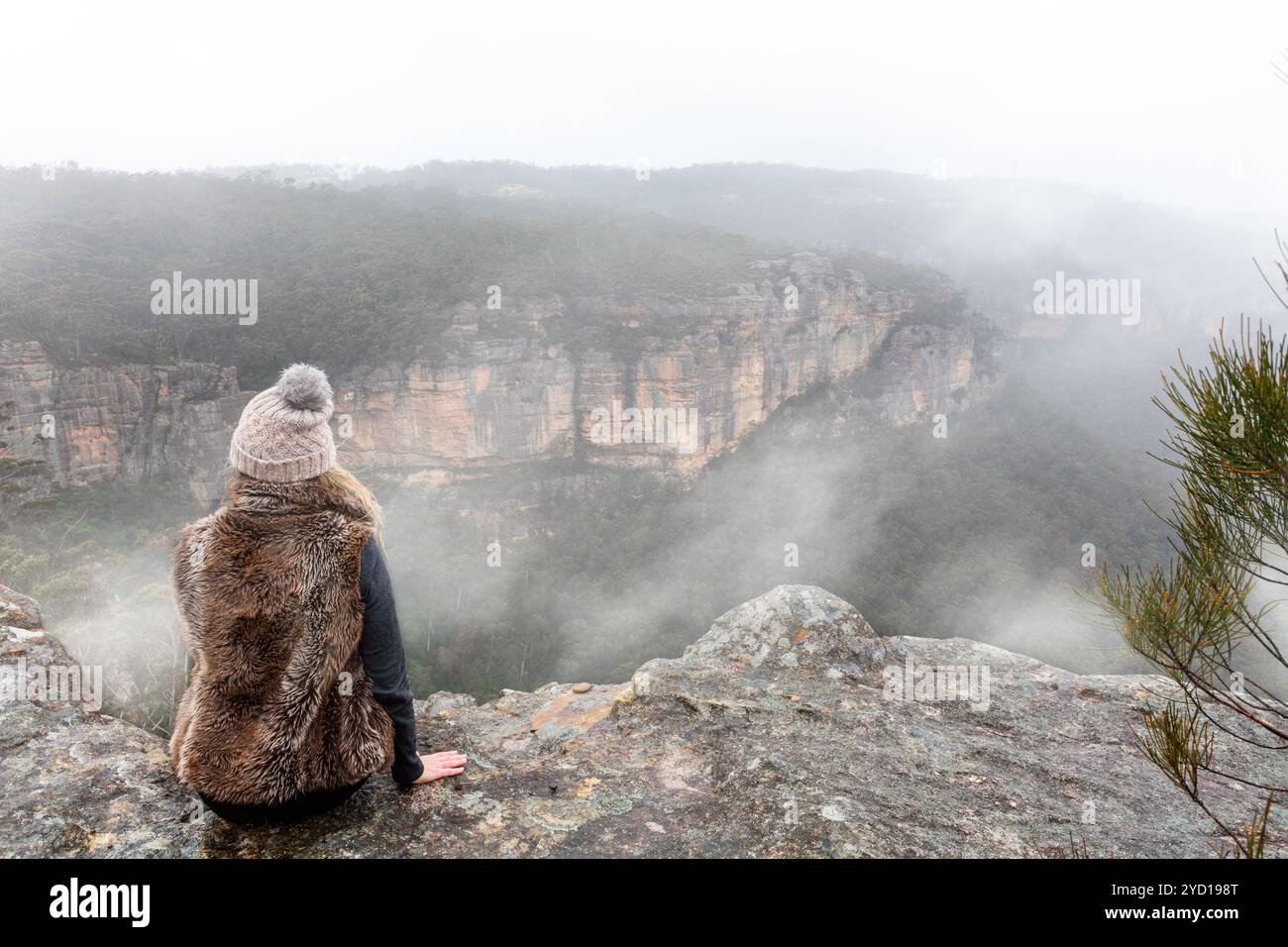 Female sitting on mountain top cliff ledge looking out into the misty ...