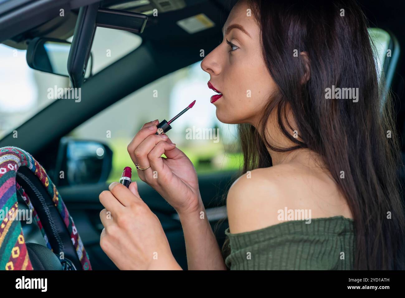 A beautiful young brunette applies makeup in her car’s mirror, getting ...