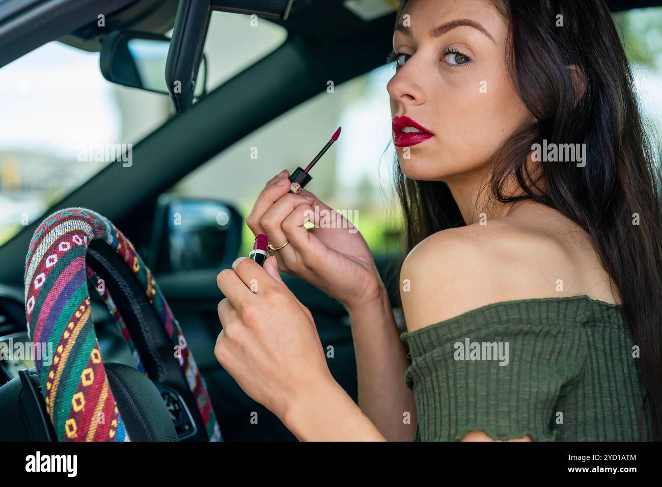 A beautiful young brunette applies makeup in her car’s mirror, getting ...