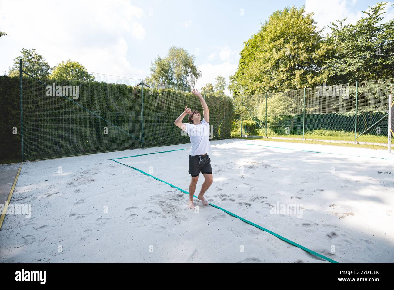 Back view of a young man serving a volleyball on a sand court under a ...