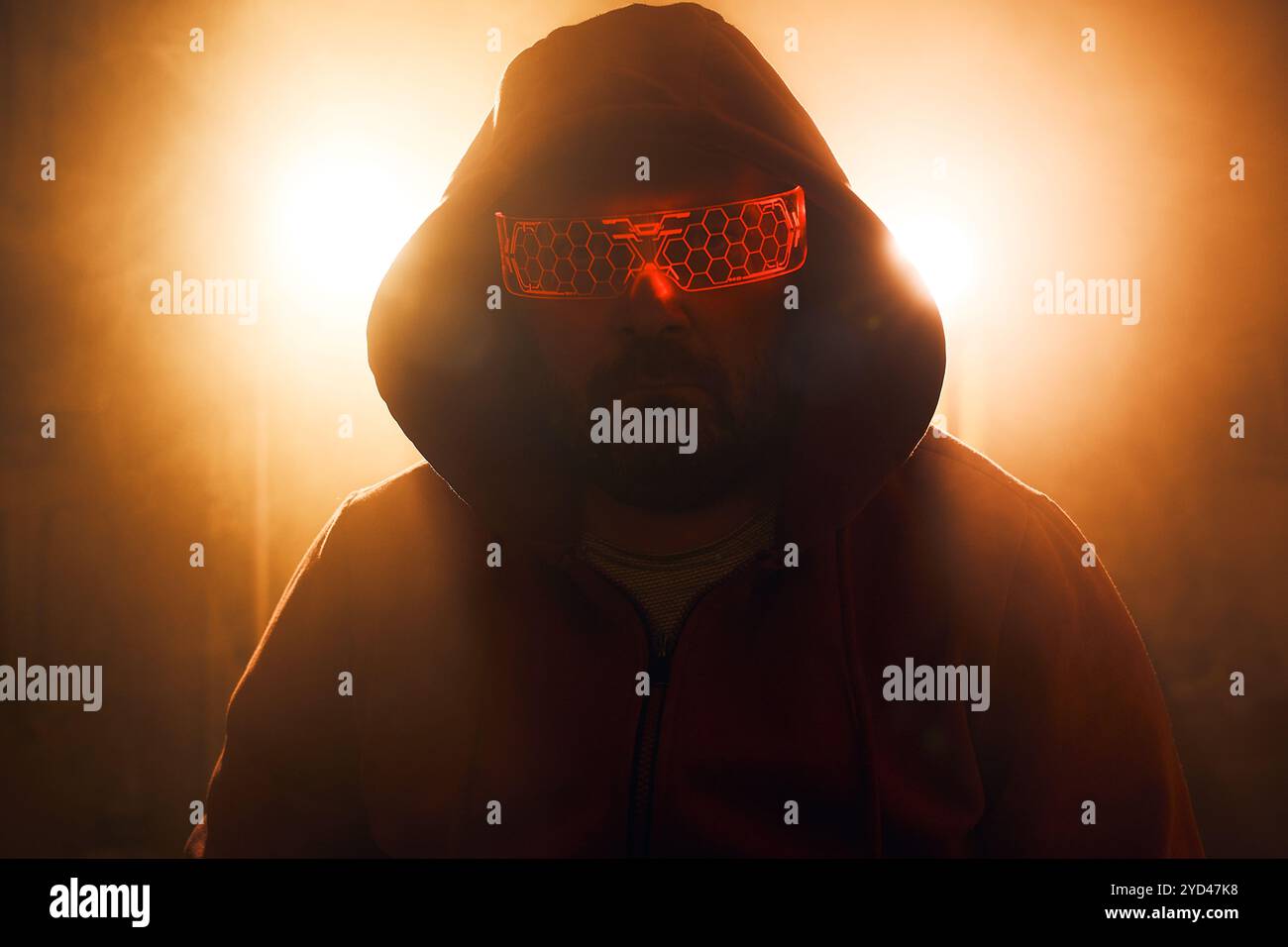 A hooded figure wearing unique glasses stands illuminated by dramatic ...