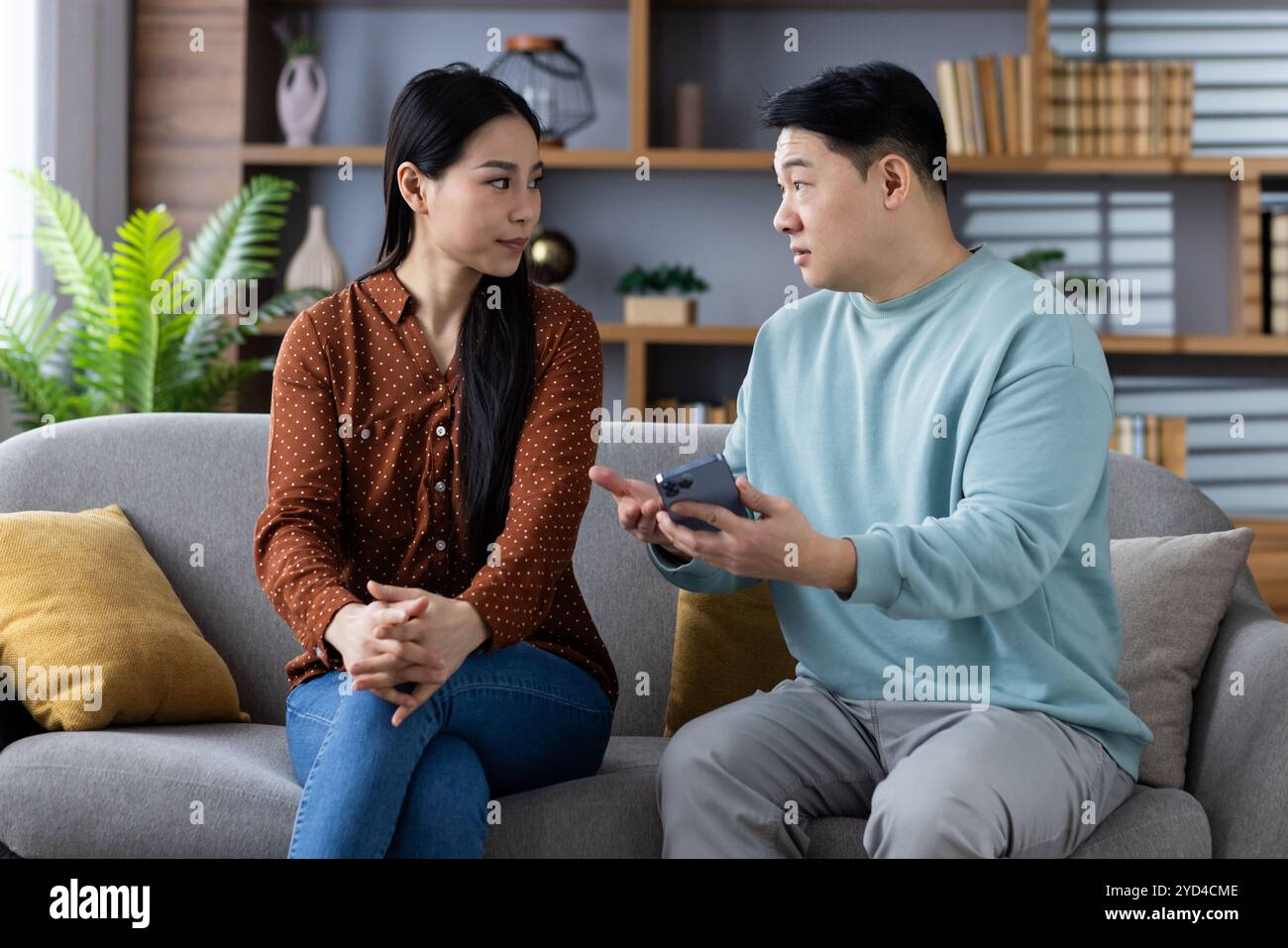 Asian couple on sofa in discussion, man holding phone while talking to woman. Scene captures ...