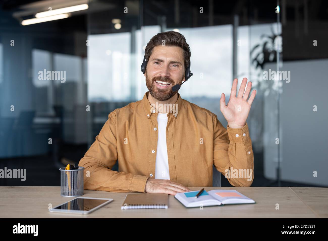 Smiling man in office wearing headset, engaged in video call, waving ...