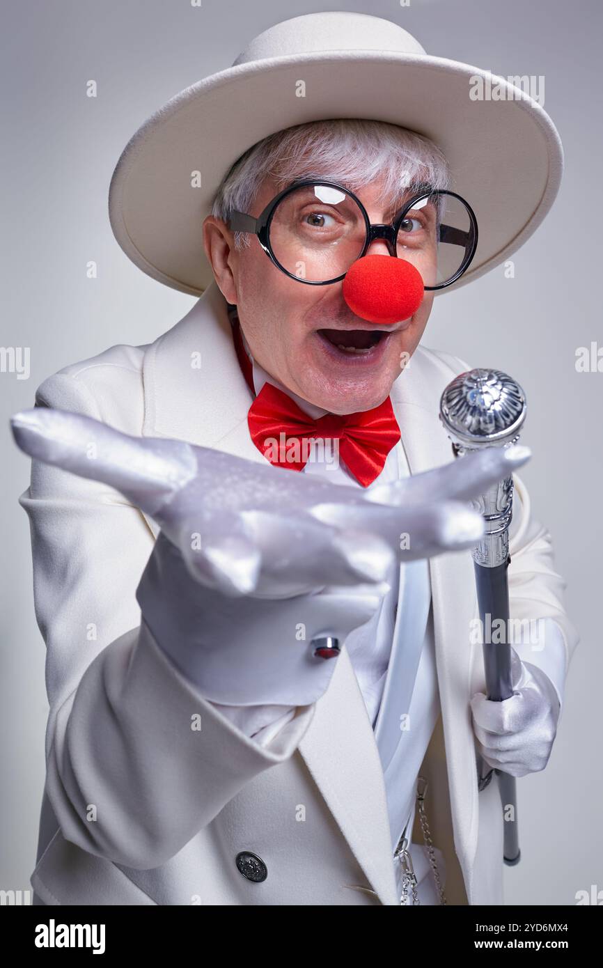 An elderly clown in a white robe and hat with a cane. The gloved hand ...