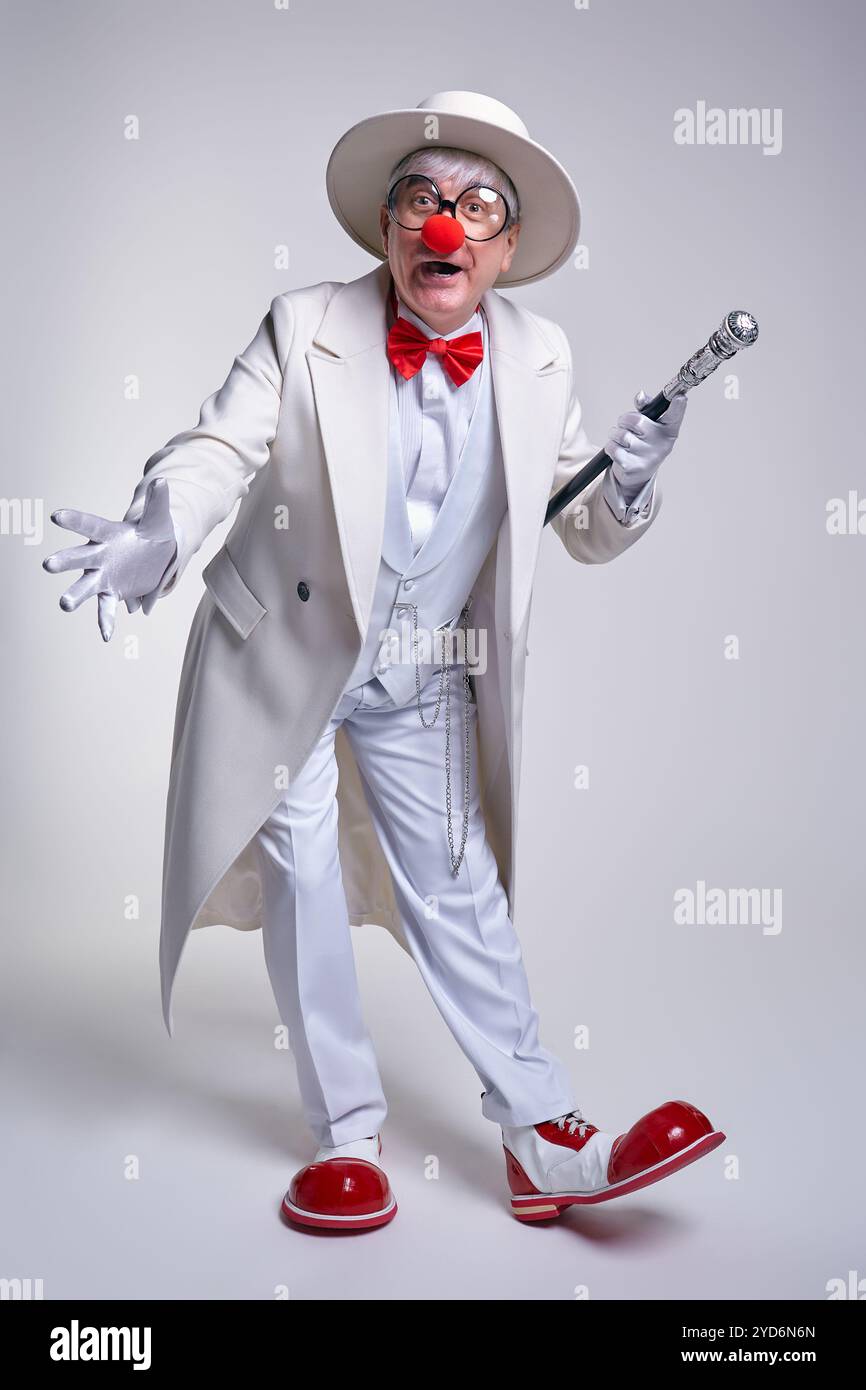 An elderly clown in a white coat, hat and large red patent leather ...