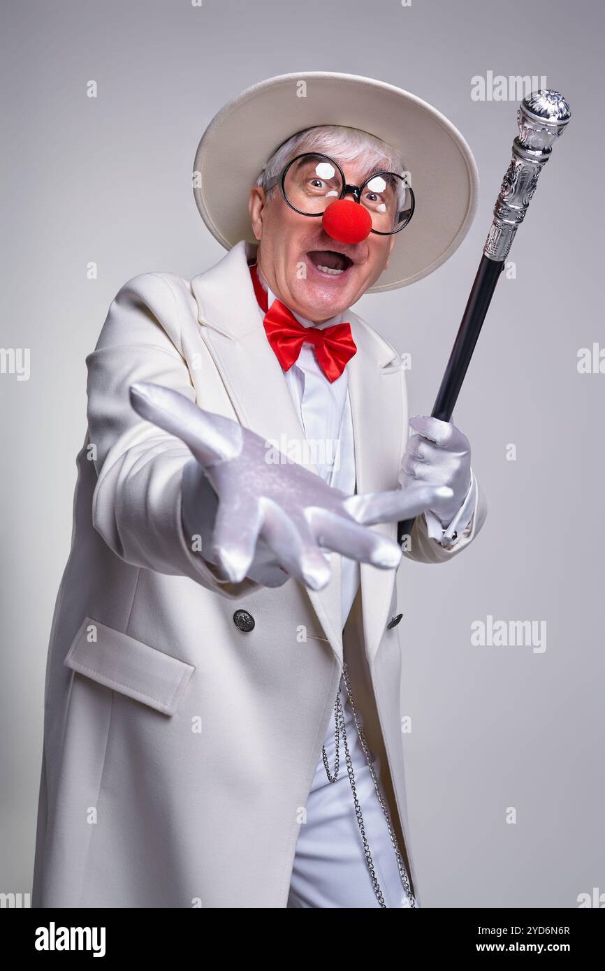 An elderly clown in a white robe and hat with a cane. The gloved hand ...