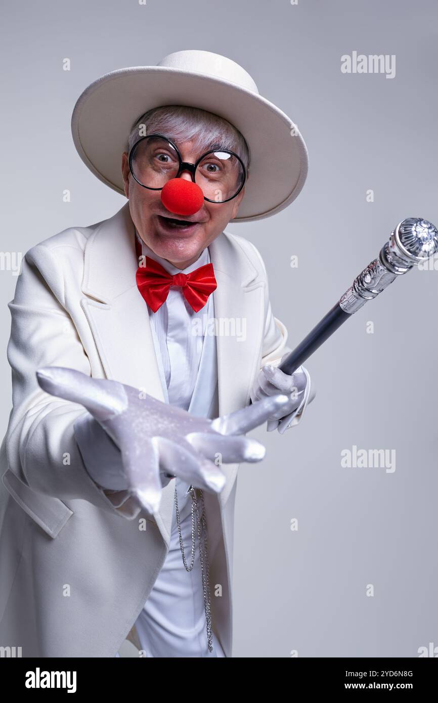 An elderly clown in a white robe and hat with a cane. The gloved hand ...