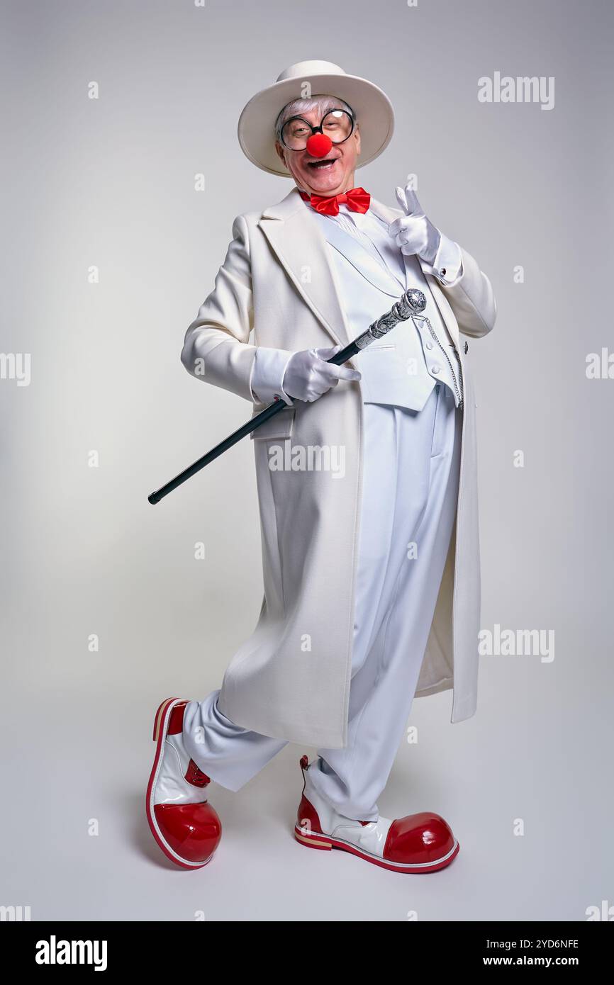 An elderly clown in a white coat, hat and large red patent leather ...