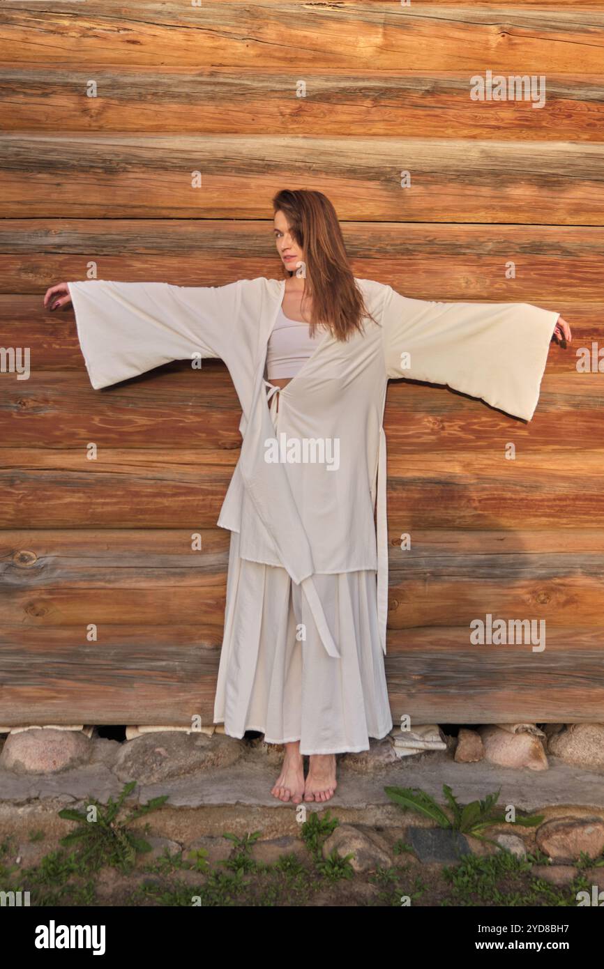 A person in flowing white garments stands by a wooden cabin, barefoot ...
