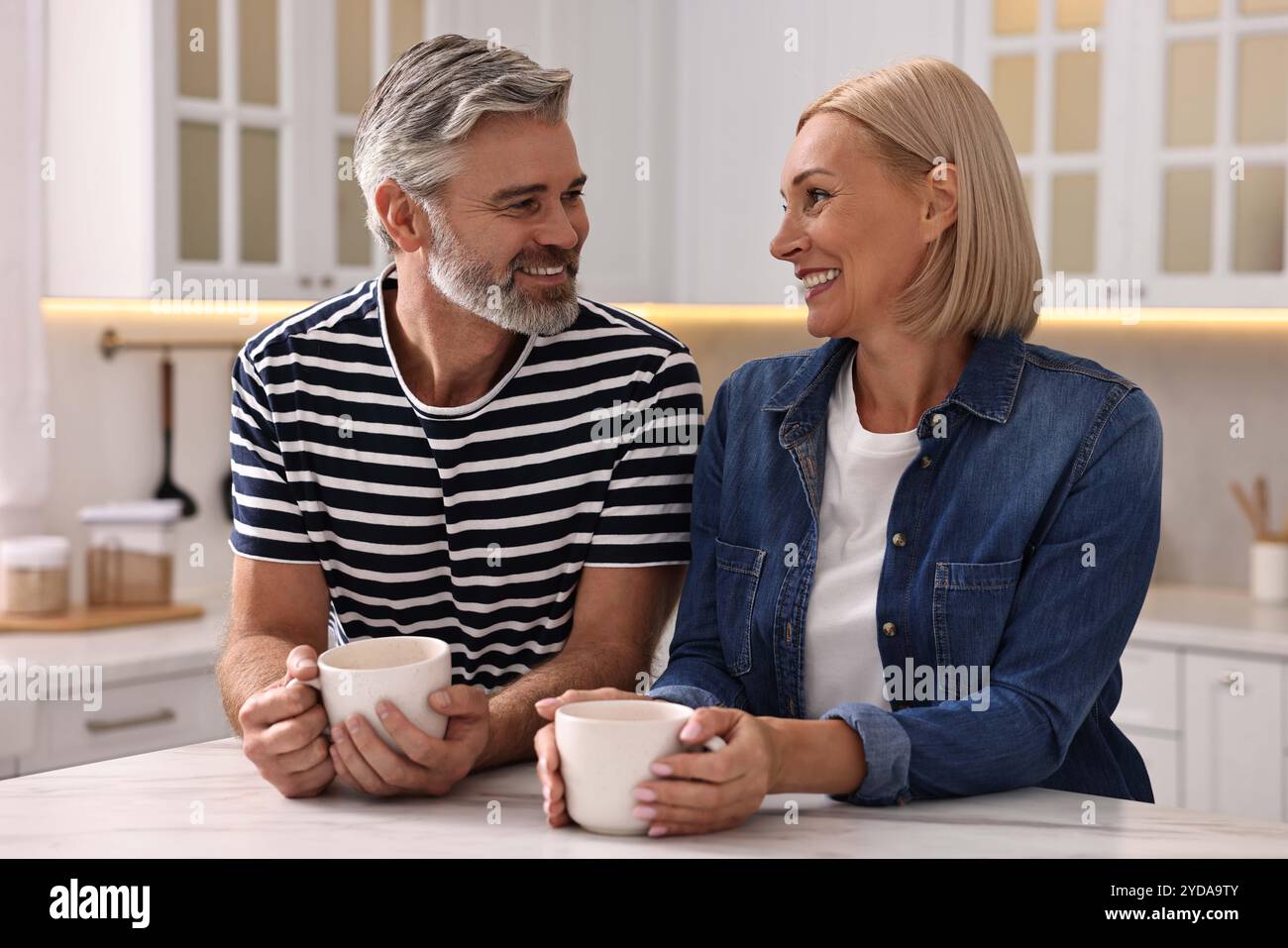 Happy middle aged couple with cups of drink at white marble table in ...