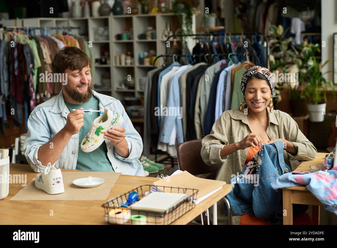 Happy young man and woman sitting by tables in thrift shop and upcycling used denim jacket and ...