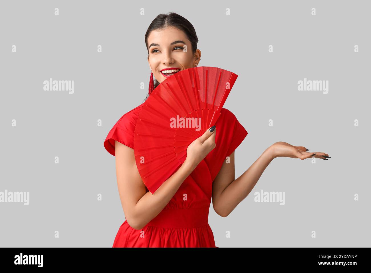 Beautiful young happy woman with fan pointing at something on grey ...