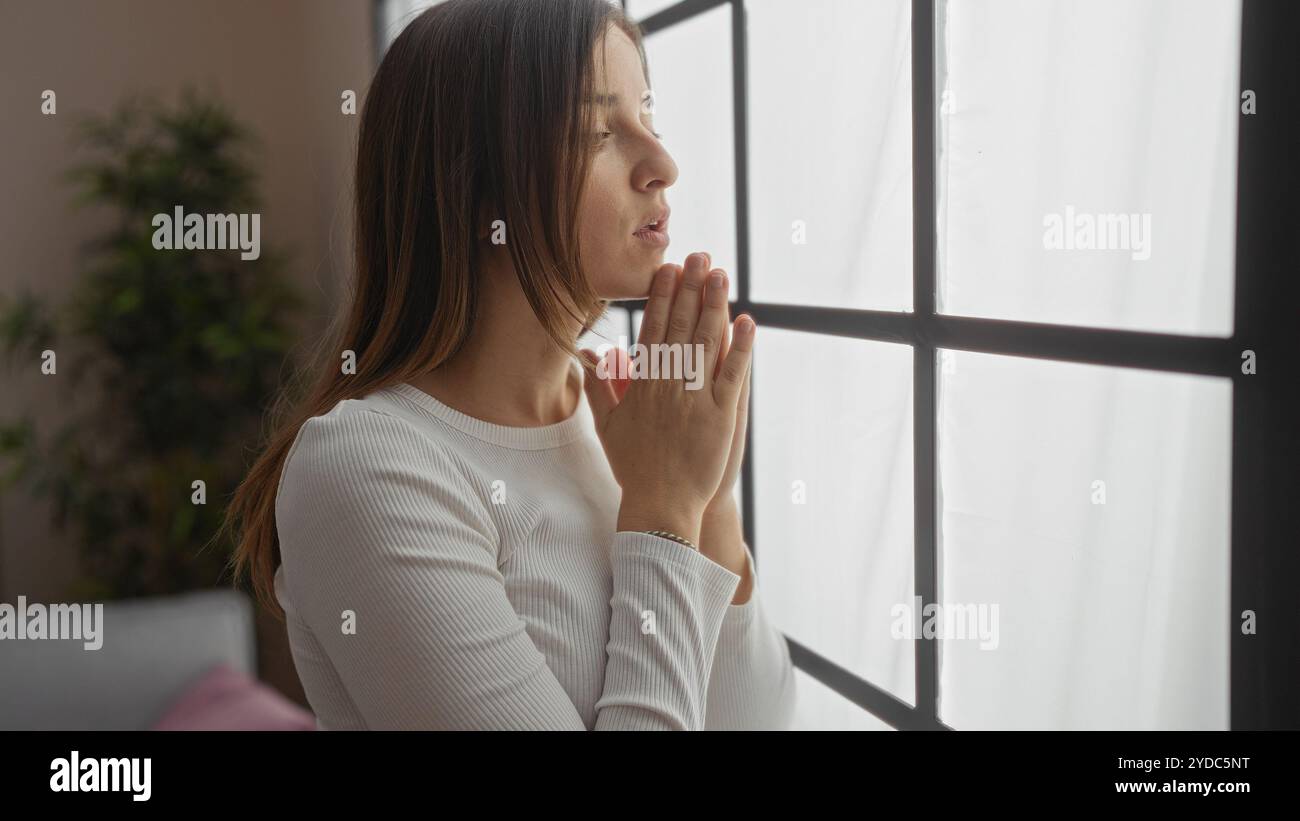 A young woman stands indoors by a window with hands clasped in a prayer ...