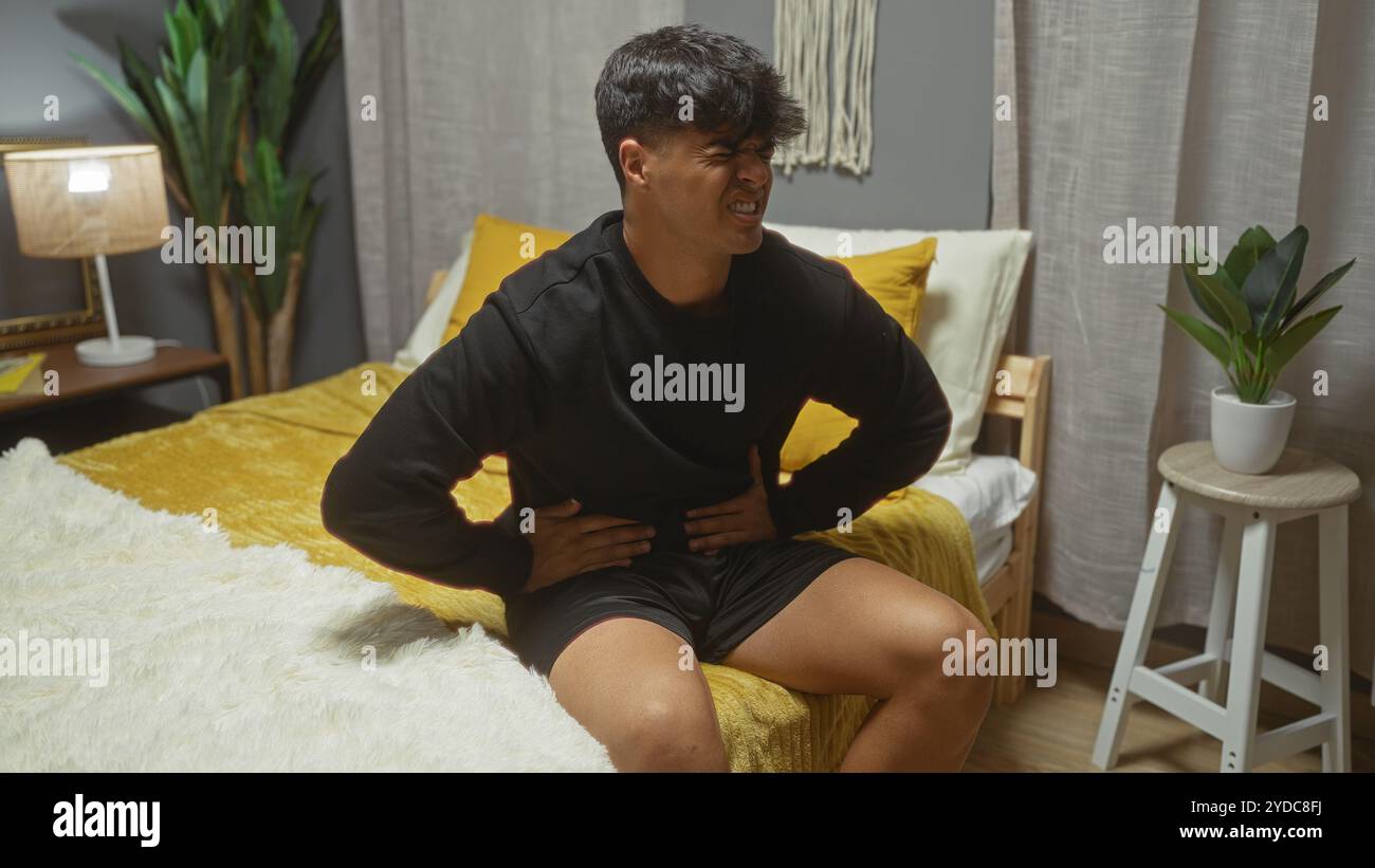 A handsome young hispanic man experiencing abdominal pain, sitting on a bed in a cozy bedroom ...
