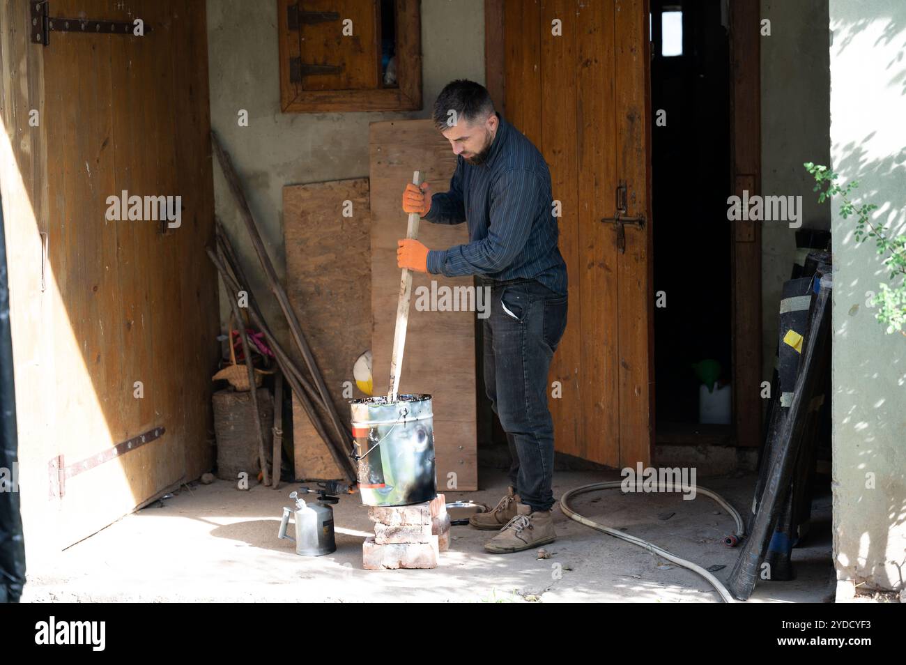 A man stirs liquid bitumen in a bucket with a wooden stick ...