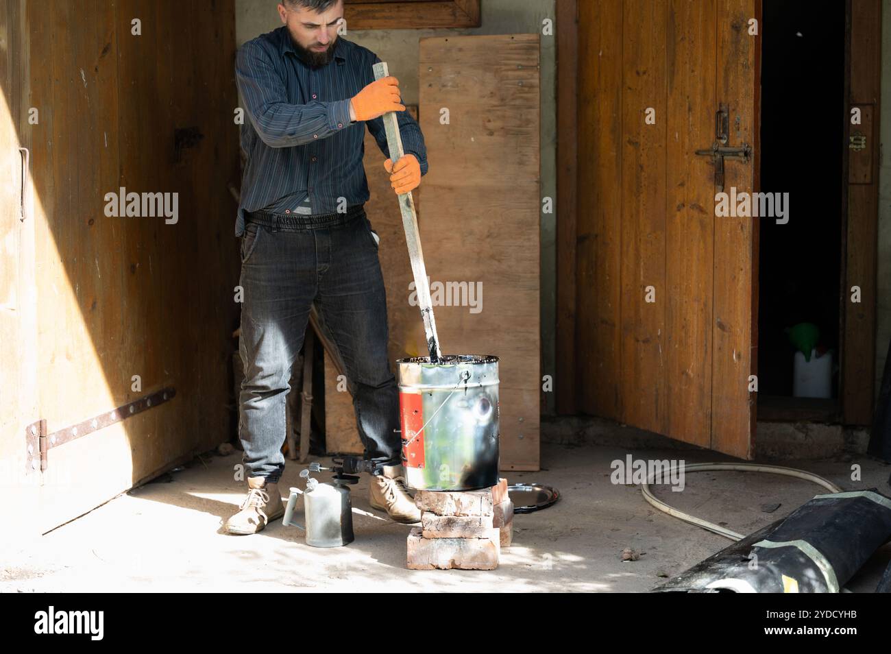 A man stirs liquid bitumen in a bucket with a wooden stick ...