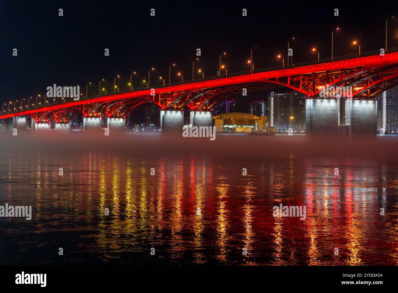 A red illuminated bridge stretches over a calm river at night, with ...