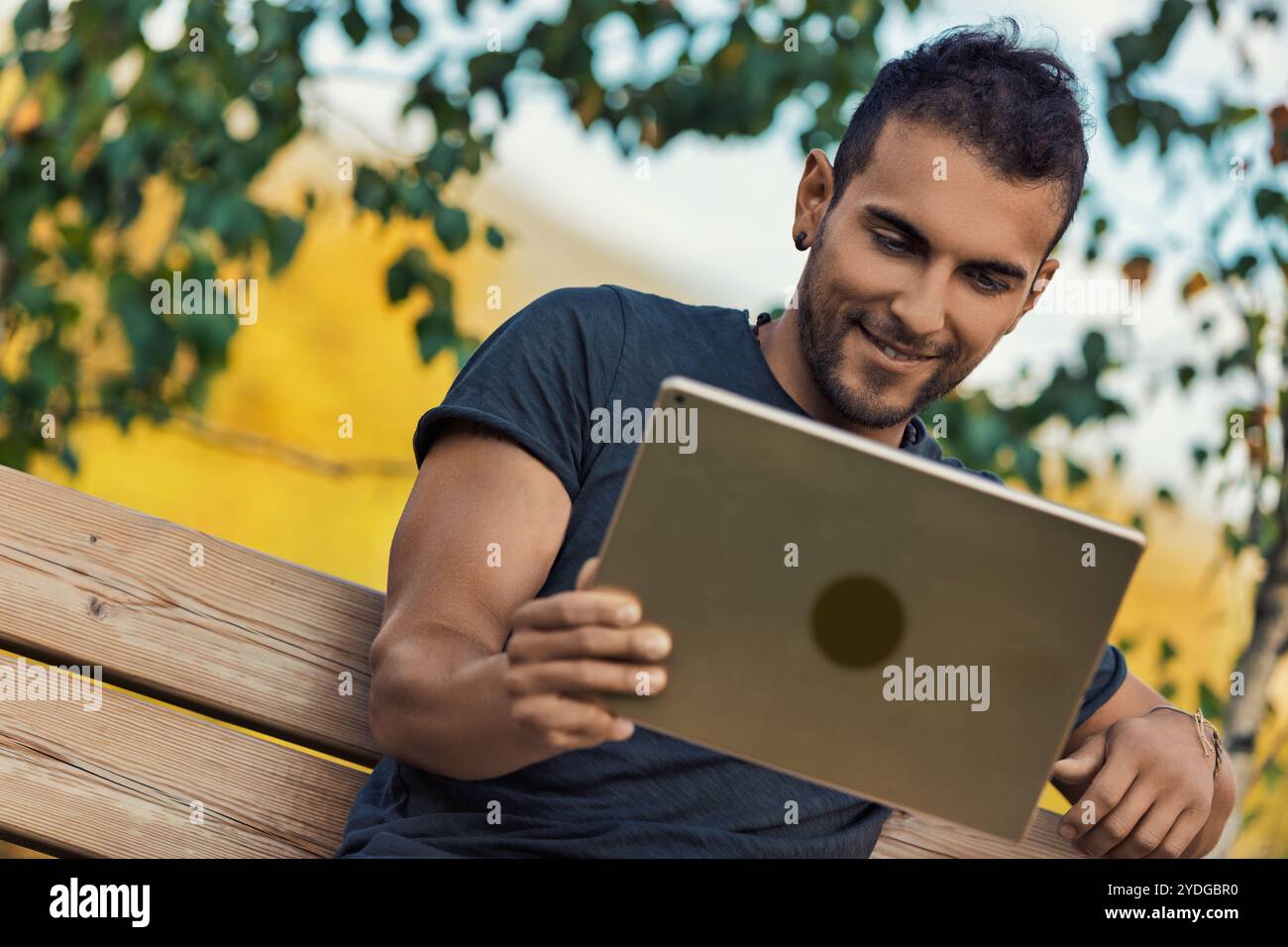 Young man enjoys using his tablet while sitting on a park bench, combining technology with ...
