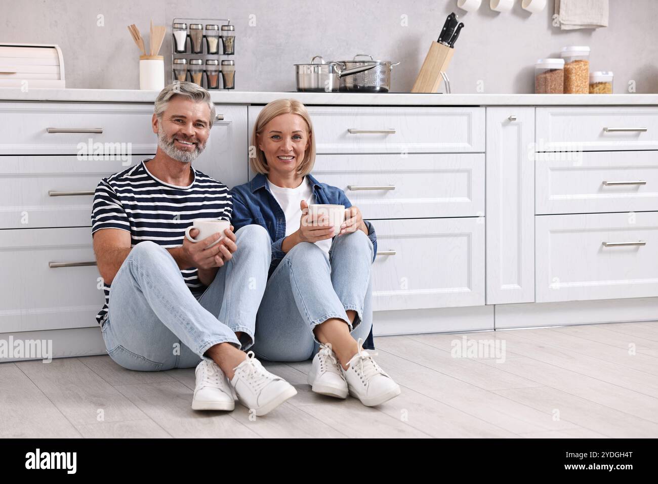 Happy middle aged couple with cups of drink on floor in kitchen, space ...