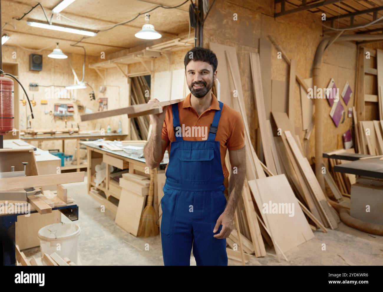 Smiling woodworker carrying timber plank on shoulder over workshop ...