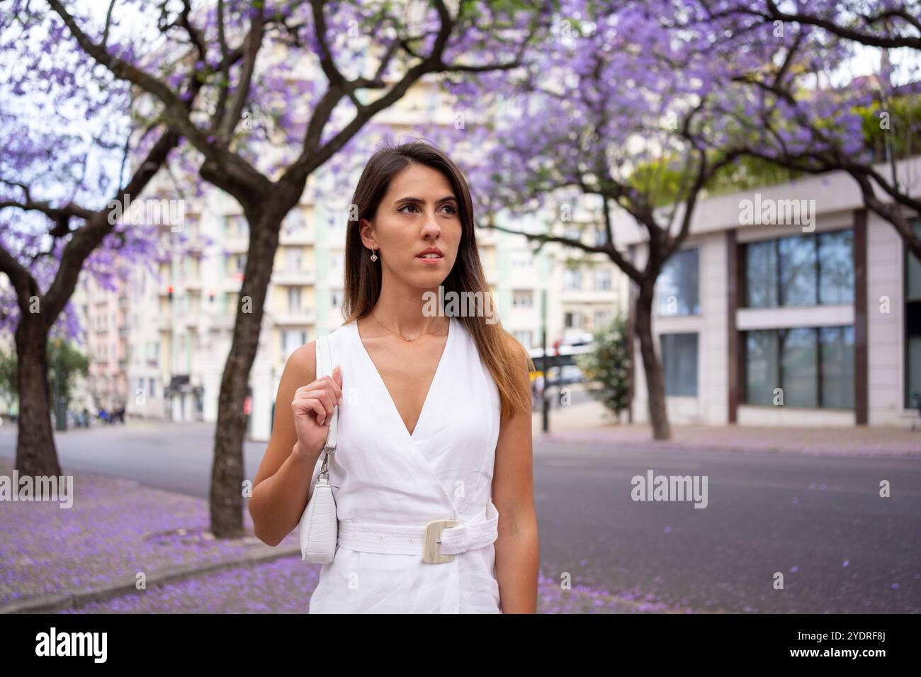 Thoughtful woman in white dress carrying purse in city during daytime. Beautiful young female is ...