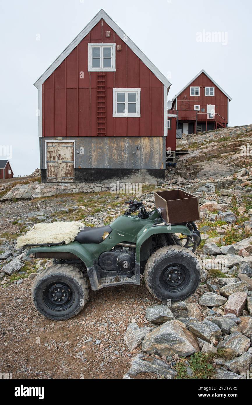 A green quad bike or ATV stands in front of red, wood-clad houses on a rocky hill, remote Arctic ...