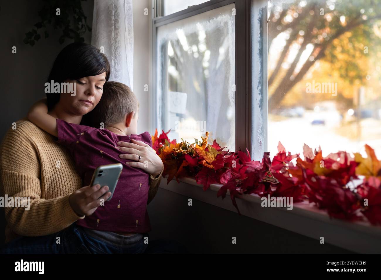 A mother holds her child by a window, phone in hand, fall background ...