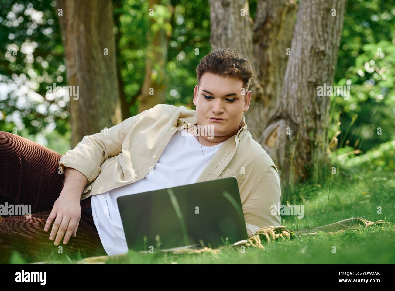 A queer person relaxes in a lush green setting while using a laptop ...