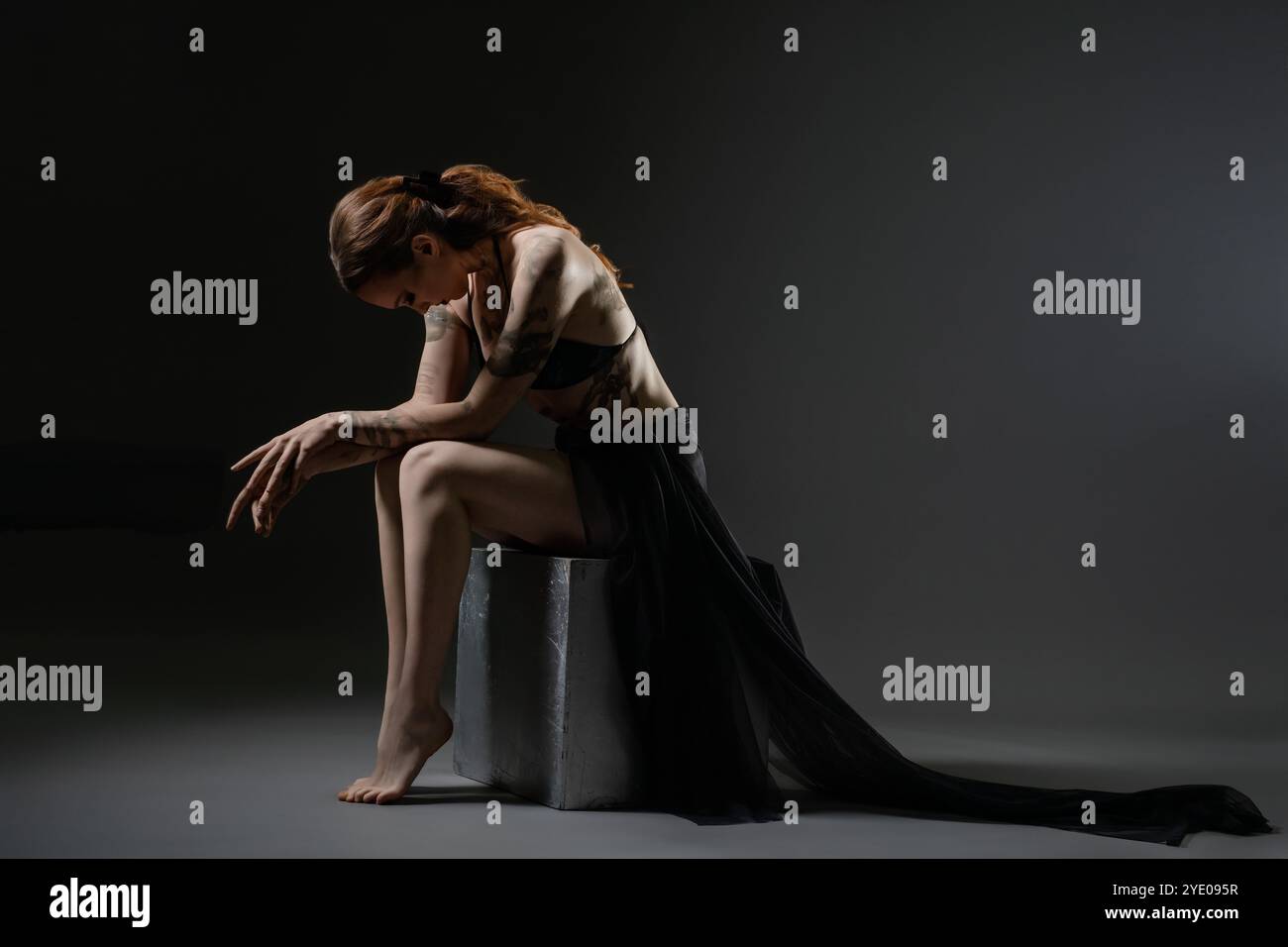 A contemplative dancer in a flowing dress poses on a stool in dim ...