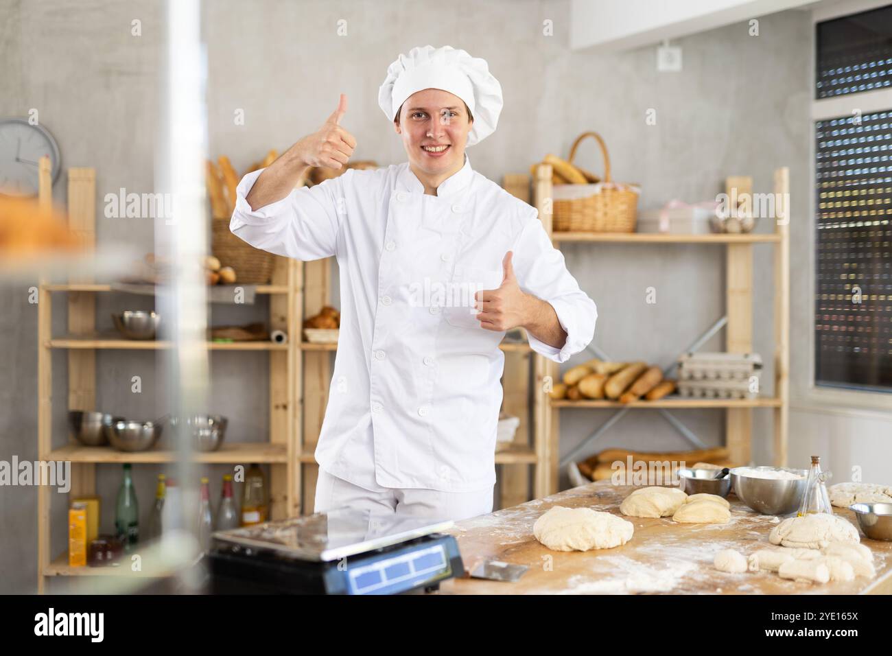 Satisfied young male baker gives thumbs up posing in own small bakery ...