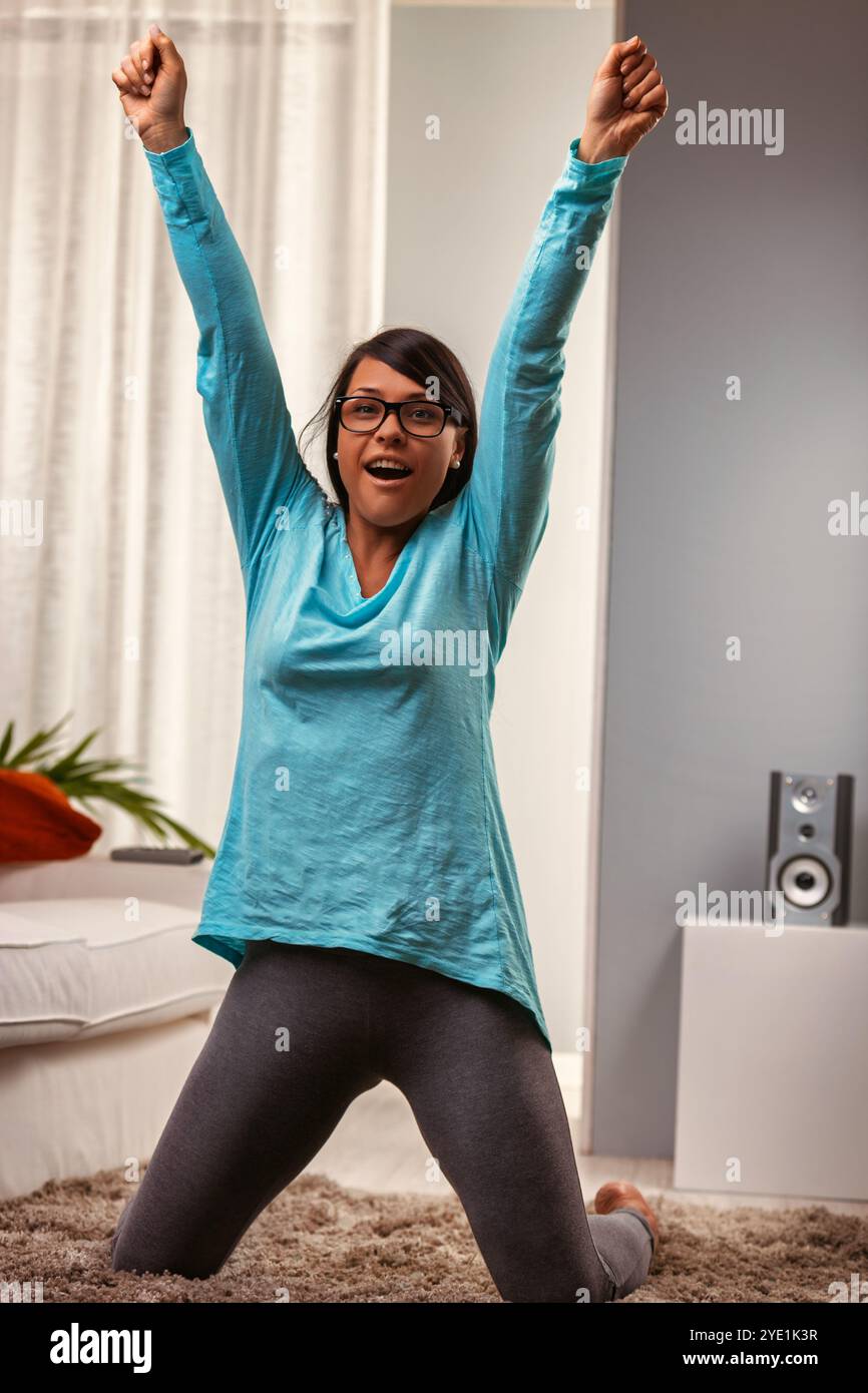 Excited young woman is kneeling on the carpet with her arms raised in a ...