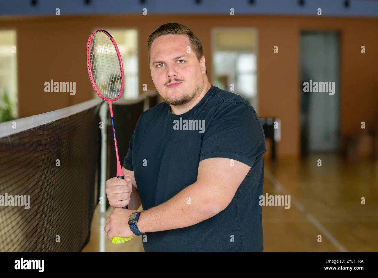 A man stands confidently on an indoor badminton court, holding a pink ...