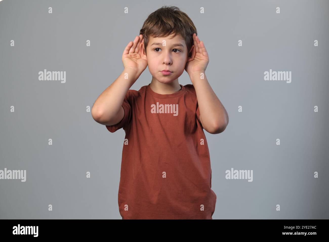 A young boy stands with hands cupped around his ears, as if listening ...