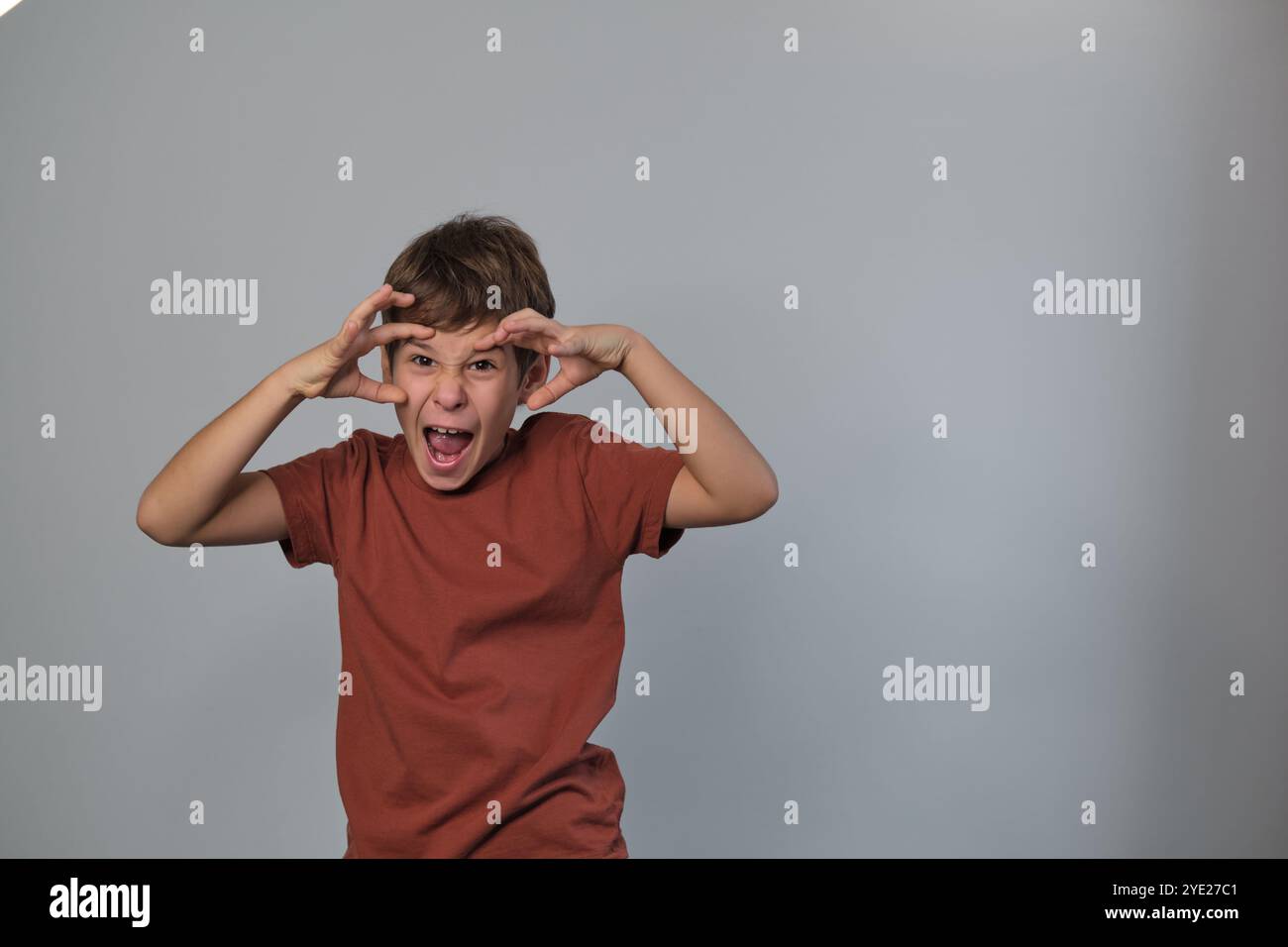 A boy in a rust-colored shirt makes a monster face, fingers pulling his ...