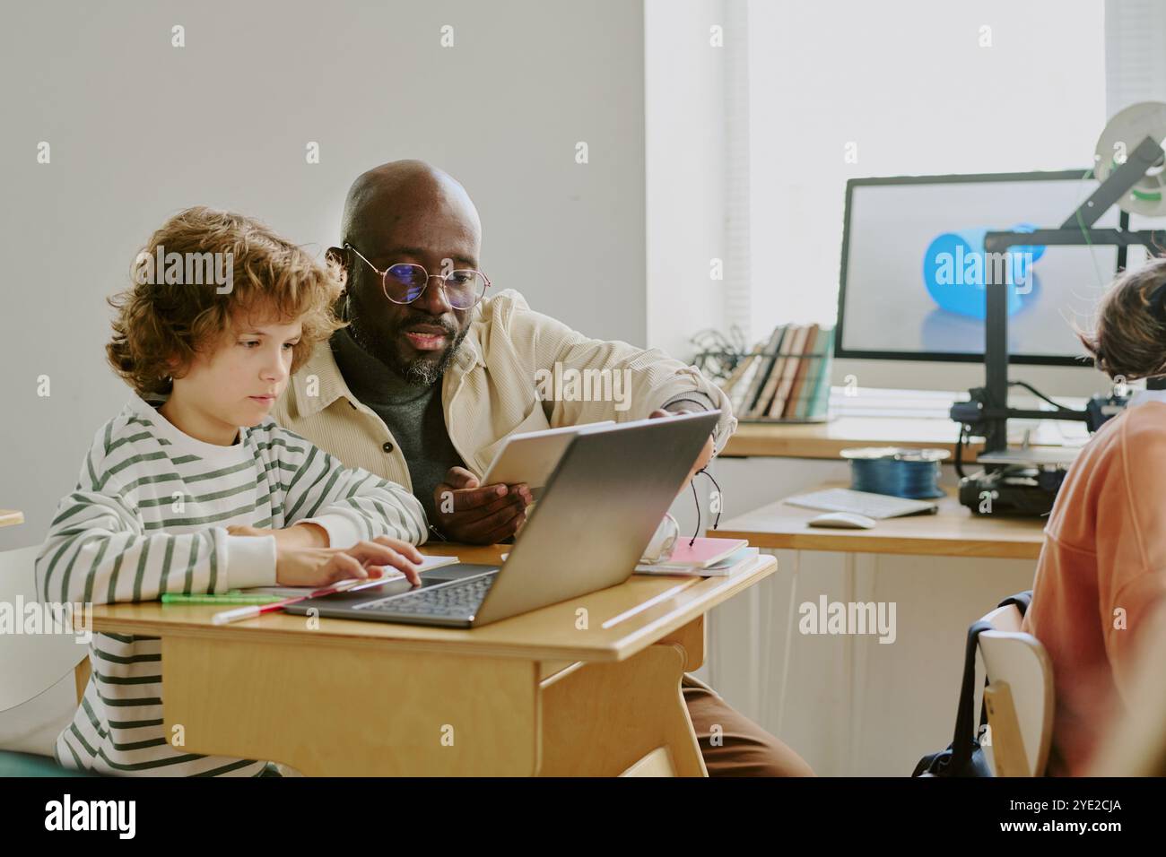 African American teacher mentoring focused young student using laptop in bright classroom. Books ...