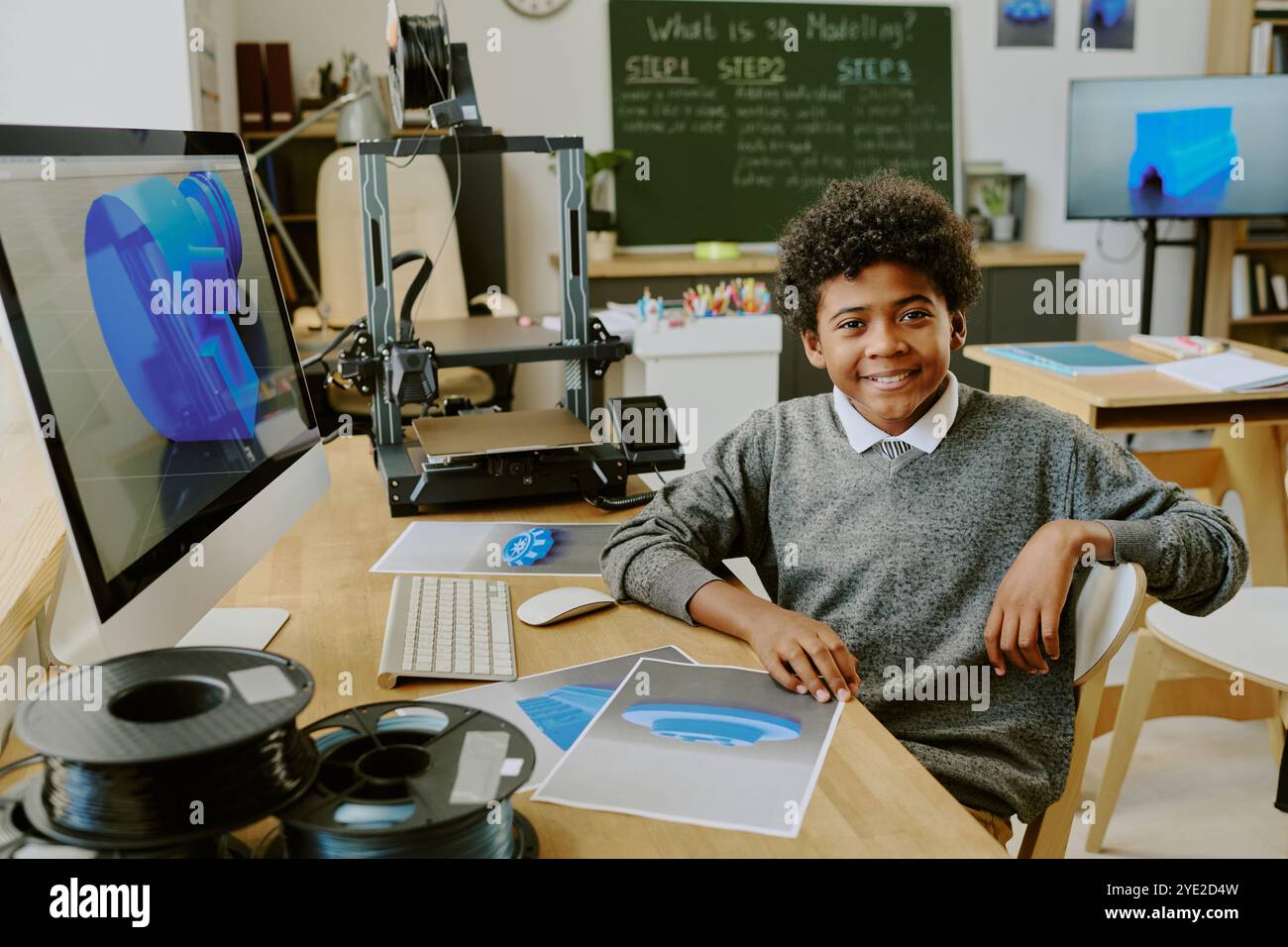 Boy smiling while sitting at desk with 3D printing equipment and ...