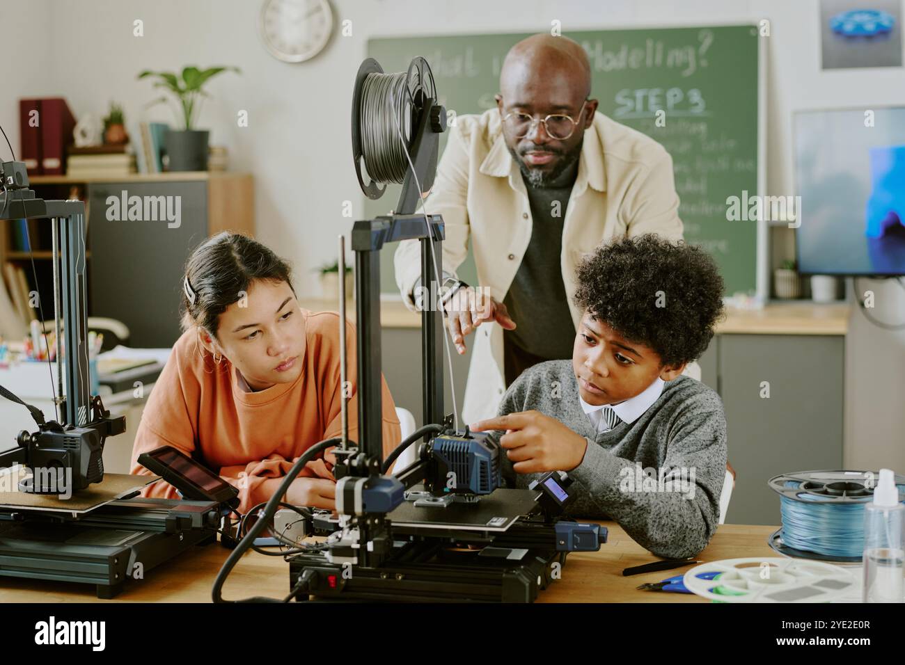 Teacher guiding two students in using 3D printing technology in classroom equipped with 3D ...